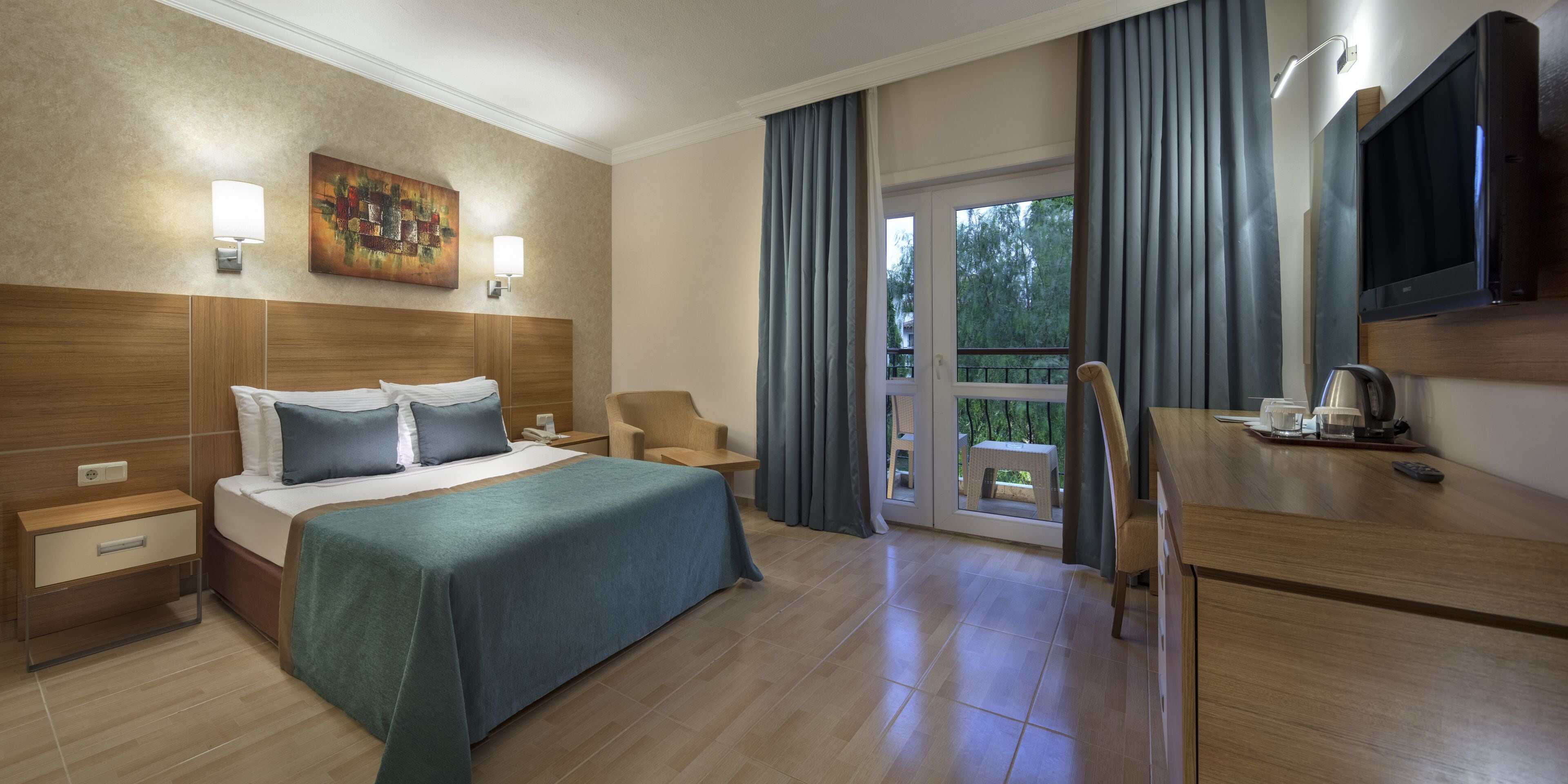 Quarto de hotel com cama, escrivaninha, cadeira, televisão e varanda com cortinas.
