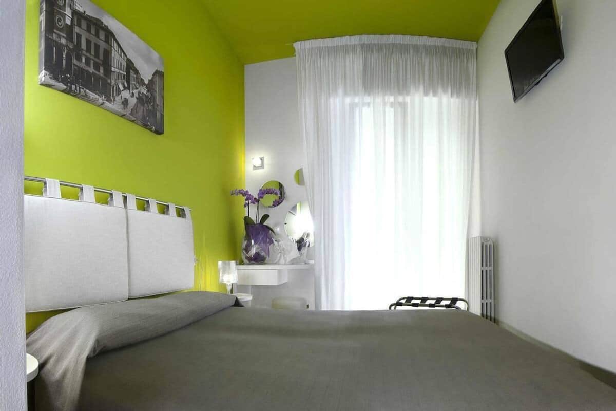 Quarto de hotel com cama, mesa de cabeceira branca, espelho, janela com cortinas brancas e televisão fixada na parede.