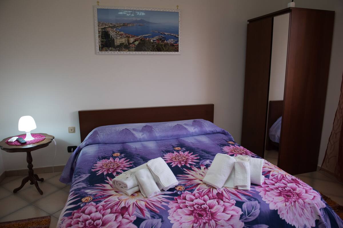 Quarto com cama, mesa de cabeceira com abajur, espelho e um quadro na parede.