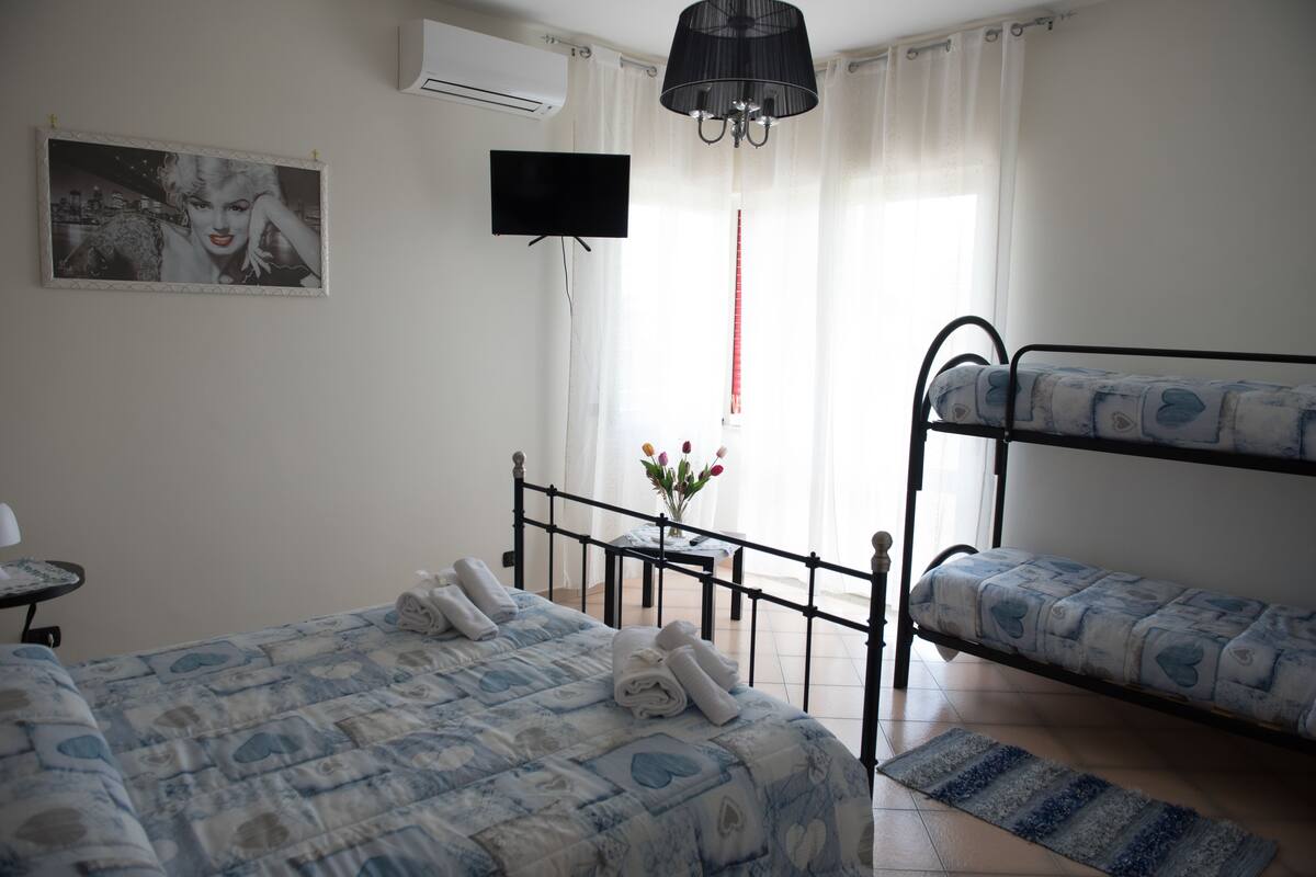 Quarto de hotel com uma cama, televisão, janela com cortinas e uma cama beliche.