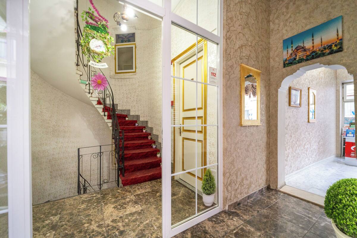 Um hall de hotel com piso de mármore, escada com carpete vermelho, placa de boas-vindas e pintura de uma mesquita.