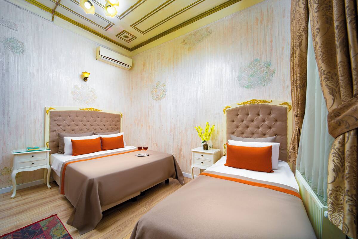 Quarto de hotel com duas camas, uma mesinha de cabeceira, ar condicionado e uma janela com cortinas.