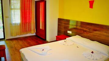 Quarto de hotel com piso de madeira, cama com lençóis e toalhas brancas, porta vermelha e parede amarela com placa de número vermelha.