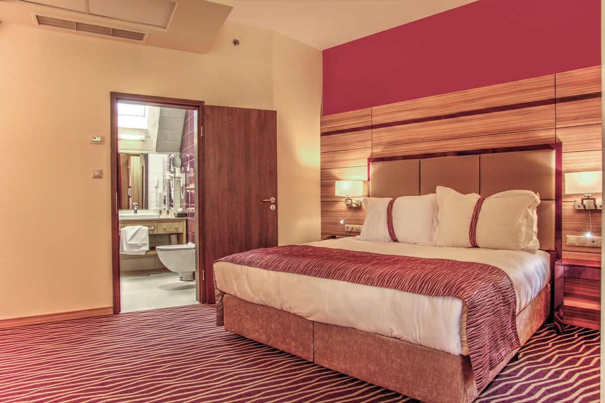 Quarto de hotel com uma cama grande, cabeceira de madeira, banheiro visível através de uma porta aberta e um tapete estampado.