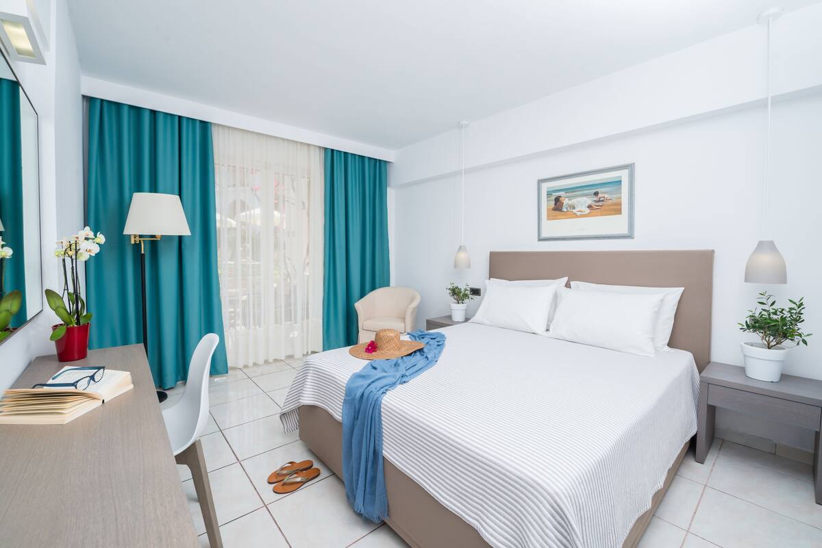 Quarto de hotel com cama, escrivaninha, cadeira e plantas em vasos.