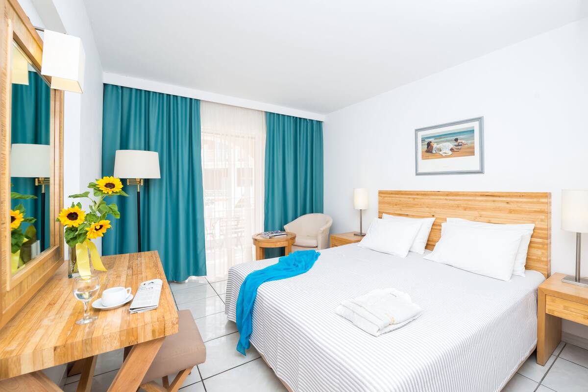 Quarto de hotel com cama, escrivaninha com cadeira, luminária, espelho e janela com cortinas.
