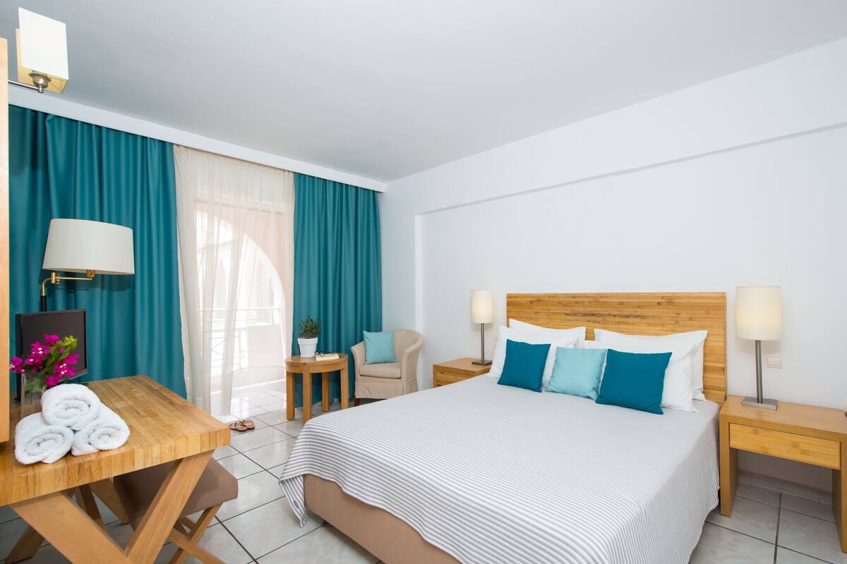 Quarto de hotel com uma cama grande, duas mesas de cabeceira, uma escrivaninha de madeira com toalhas dobradas, uma cadeira, televisão e uma janela com cortinas.