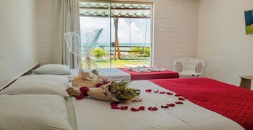 Quarto de hotel com duas camas, uma com colcha vermelha e outra com colcha branca. Há uma varanda com vista para o mar e vegetação.