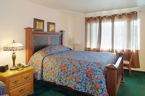 Quarto com colcha florida, cómoda de madeira com gavetas, telefone e luminária.