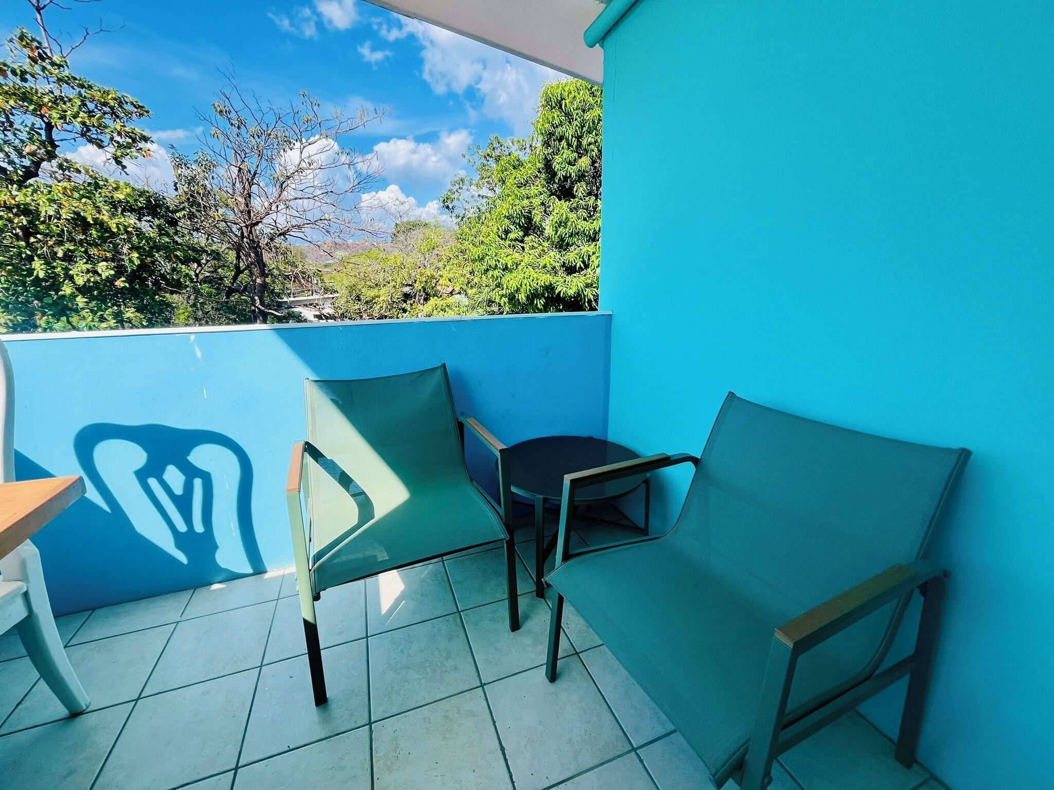Uma varanda com parede azul, uma mesa, duas cadeiras e vista para árvores e céu.