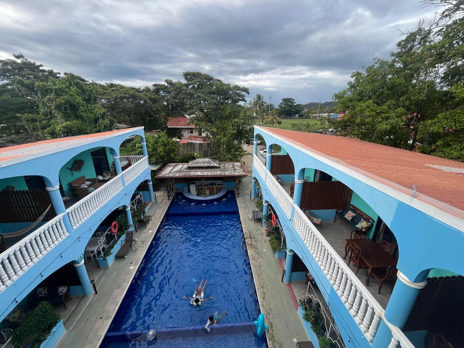 Uma piscina com dois edifícios azuis em cada lado, uma área coberta no centro e vista para o verde ao redor.