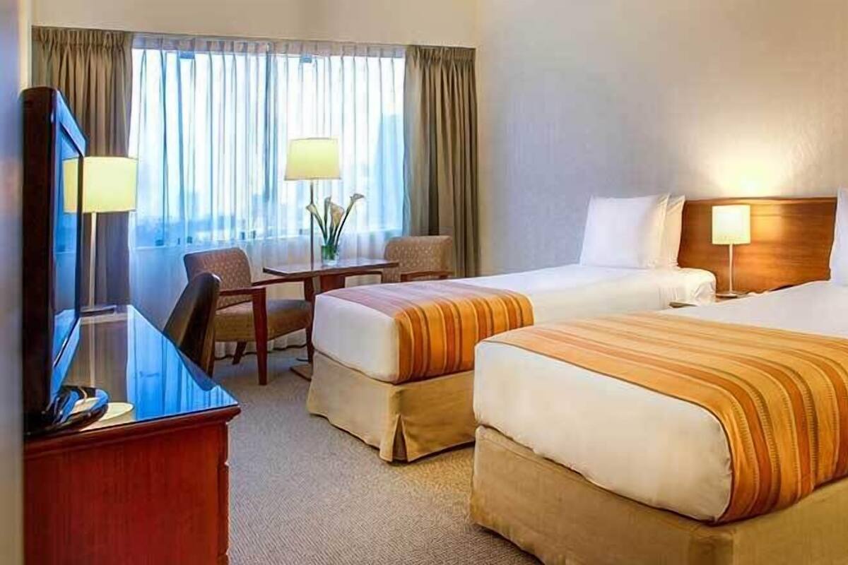Quarto de hotel com duas camas, uma mesa, uma cadeira e uma luminária.