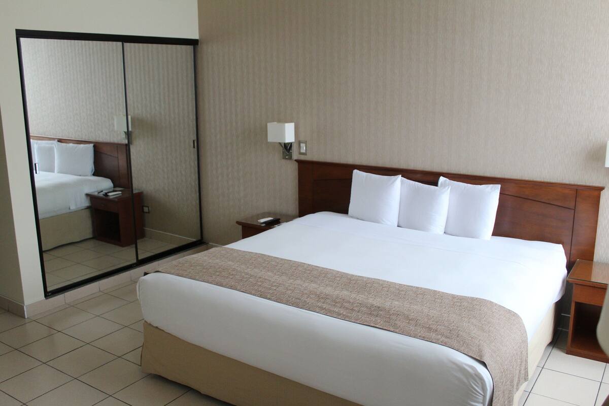 Quarto de hotel com uma cama grande, mesa de cabeceira, espelho e luminária na parede.