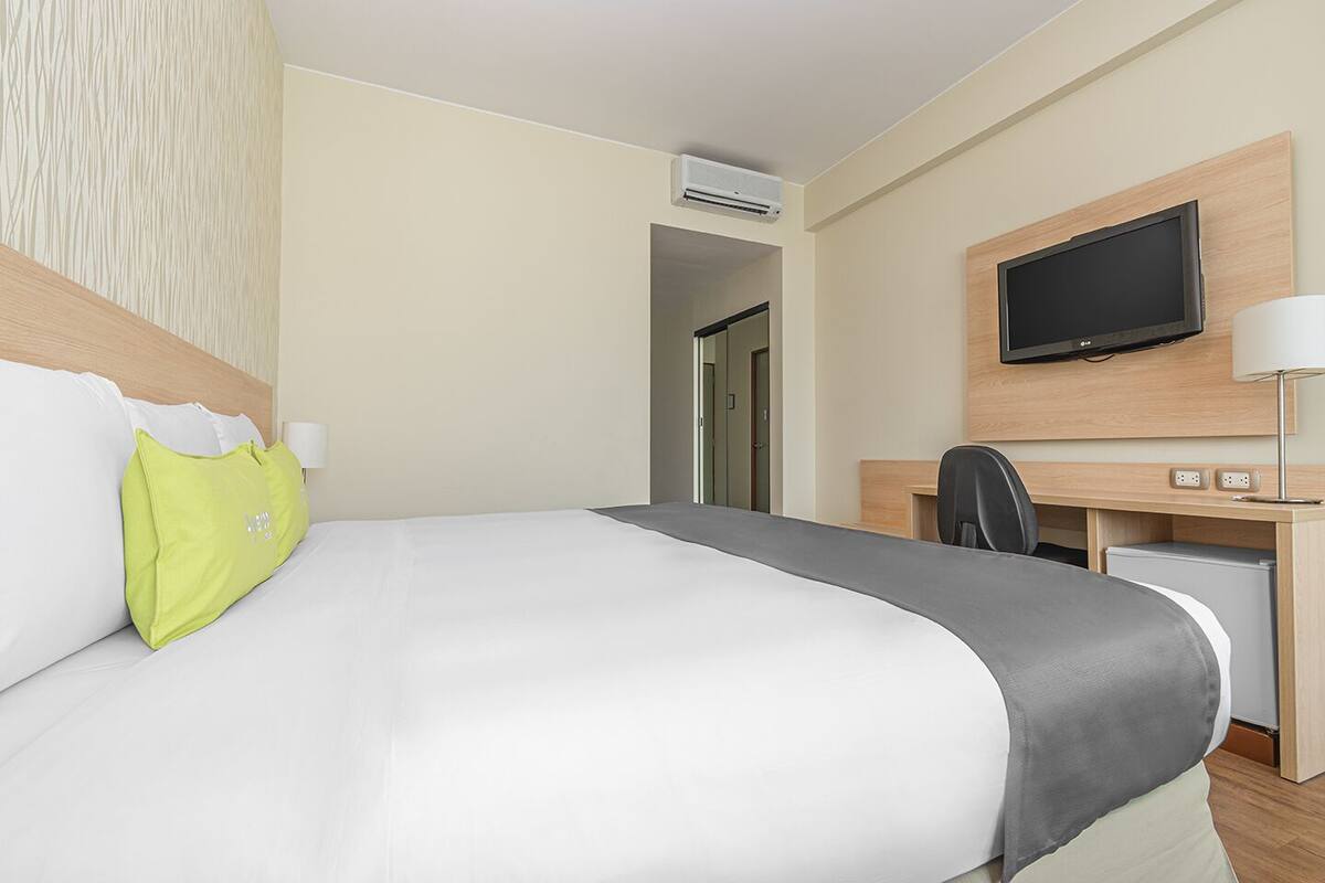 Quarto de hotel com cama, escrivaninha, cadeira, televisão e ar condicionado.