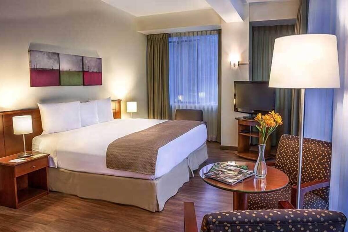 Quarto de hotel com uma cama grande, mesas de cabeceira com abajures, uma televisão, uma mesinha com um vaso de flores e uma cadeira.
