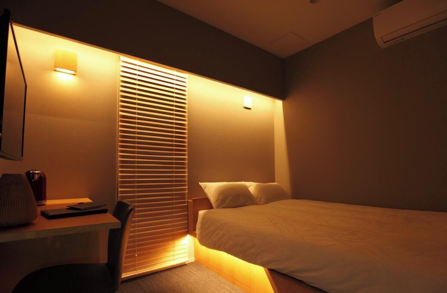 Quarto de hotel com cama, escrivaninha com luminária, cadeira e janela com persianas.