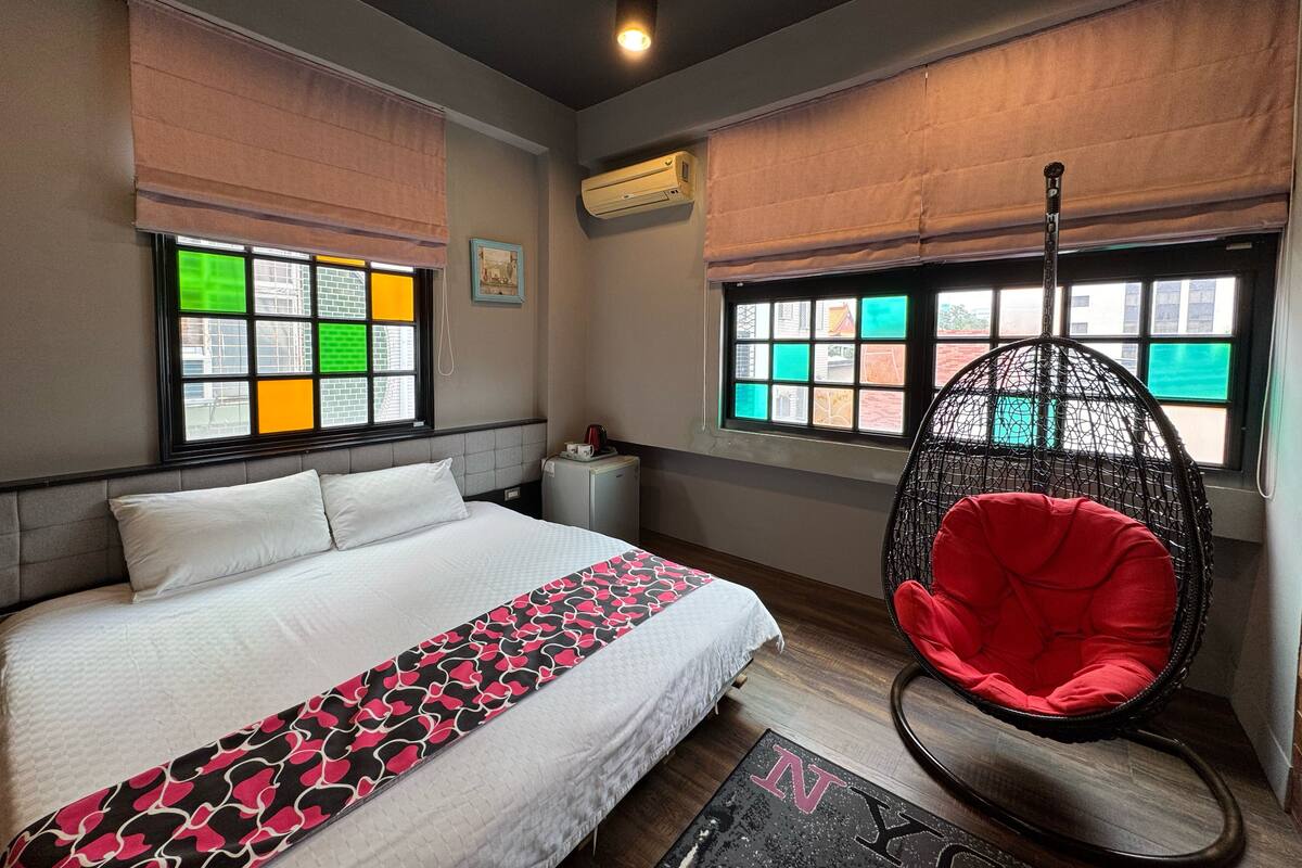 Quarto com cama, uma cadeira suspensa vermelha e duas janelas com vistas coloridas.