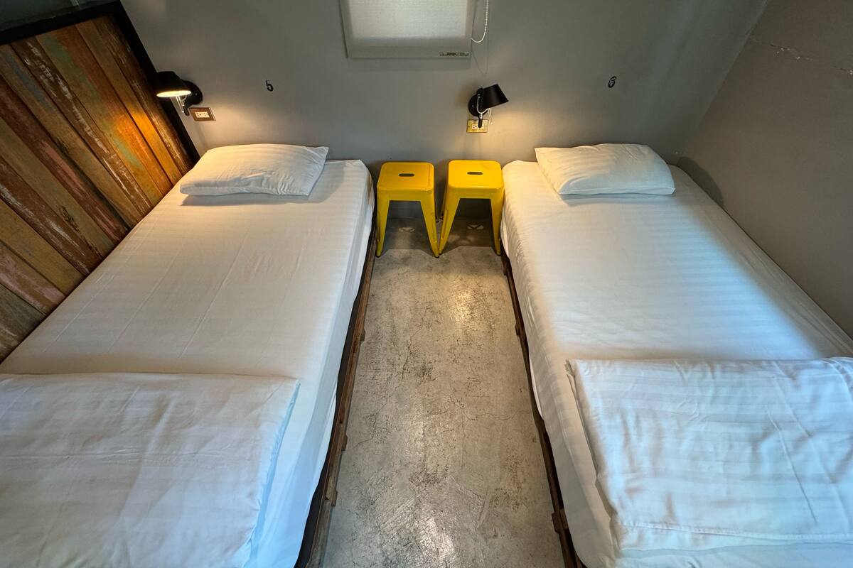 Quarto com duas camas de solteiro, cabeceira de madeira, uma pequena janela e luminária fixada na parede.