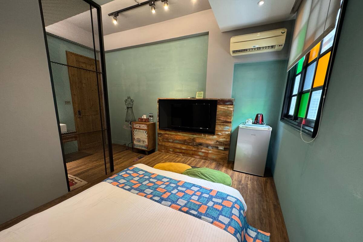 Quarto com uma cama, televisão, pequeno frigorífico e janela com vista colorida.
