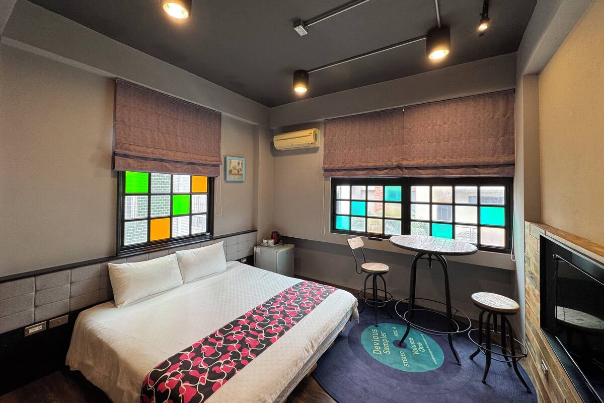 Quarto de hotel com uma cama, uma mesa pequena, dois banquinhos, uma janela com vidros coloridos e um lustre.