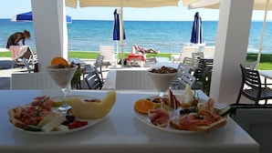 Mesa com pratos de comida e bebidas, com vista para uma praia com guarda-sóis e cadeiras.