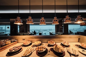 Buffet com diversos pratos, área de refeições com mesas e cadeiras, e vista panorâmica da cidade através de grandes janelas.