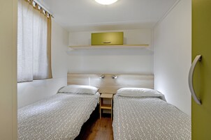 Quarto pequeno e compacto com duas camas de solteiro, uma prateleira de madeira acima das camas e uma pequena mesa no centro.