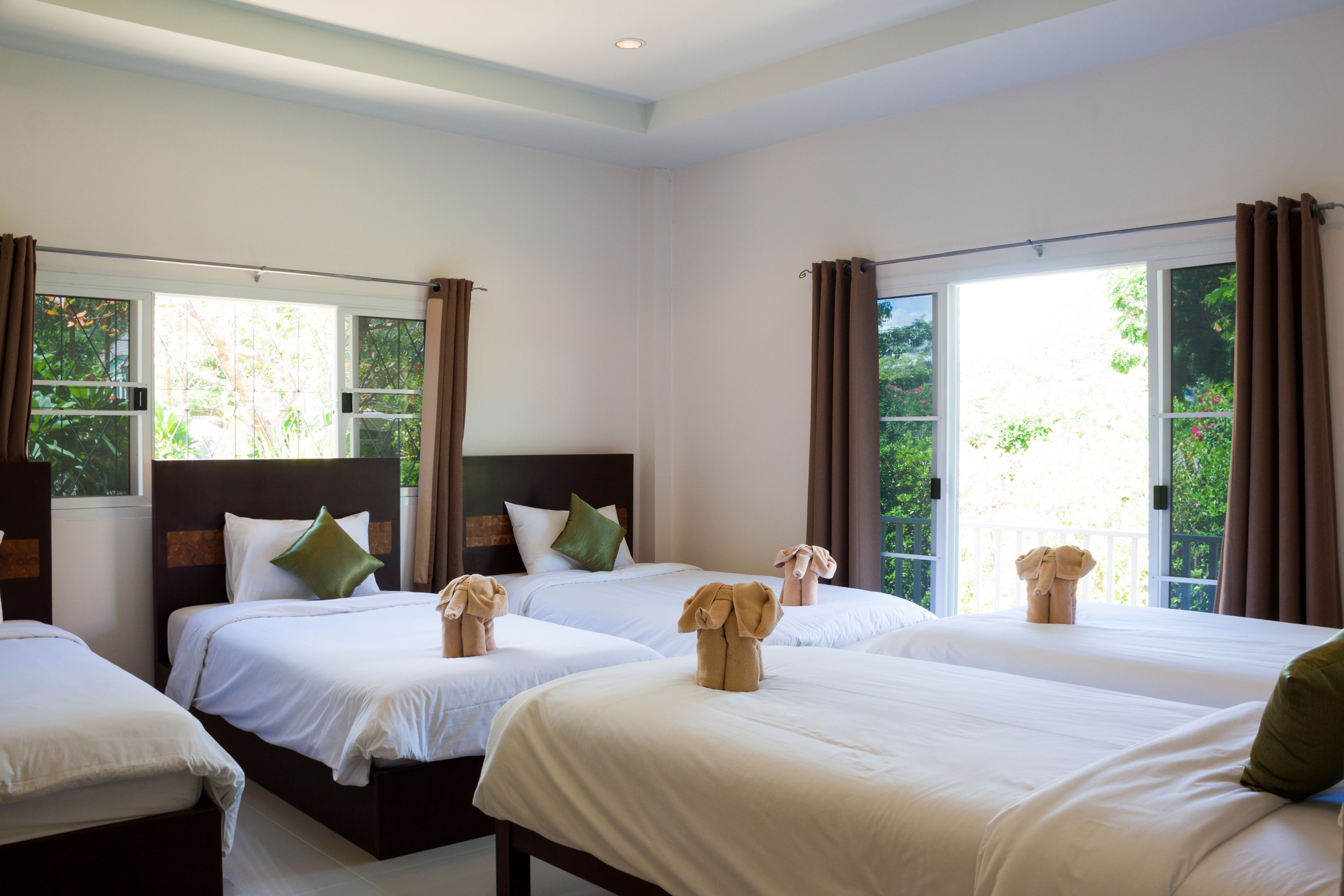 Quarto de hotel com três camas, cada uma com lençóis brancos e cabeceiras castanhas. Há cortinas nas janelas e uma pequena mesa com um vaso de flores.