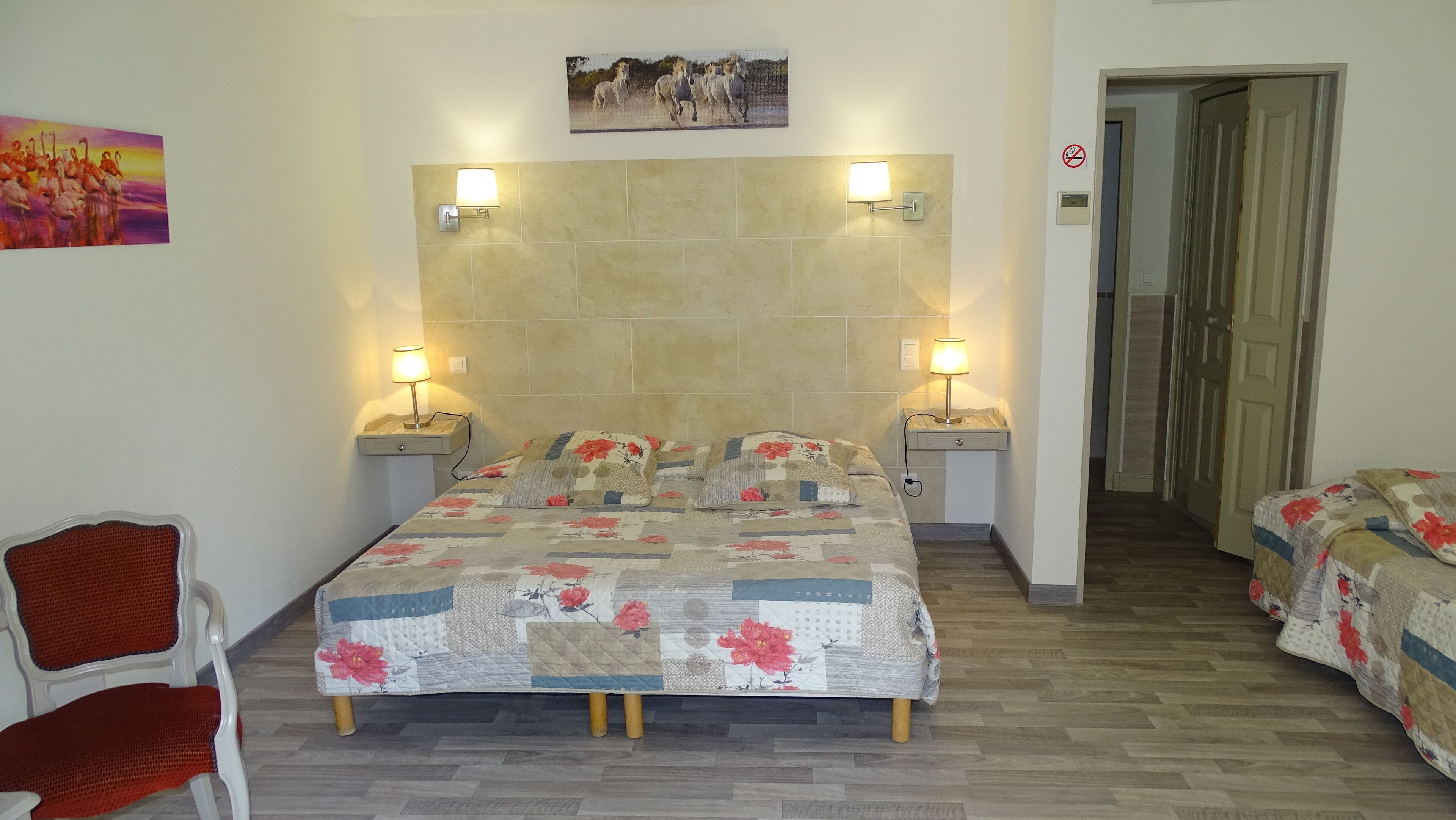 Quarto com cama, duas mesas de cabeceira com abajures, uma cadeira e uma porta que dá para outro espaço.