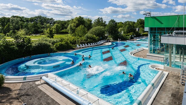 Uma grande piscina ao ar livre com várias pistas de natação e uma área de banheira de hidromassagem separada, rodeada por vegetação exuberante e um céu limpo.