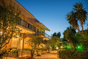 Hotel com varanda, entrada bem iluminada e jardim com palmeiras.