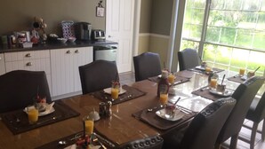 Uma sala de jantar com uma mesa grande posta para seis pessoas, cercada por cadeiras. Há jogos americanos, pratos e copos com bebidas. Uma cafeteira e outros eletrodomésticos da cozinha são visíveis ao fundo.
