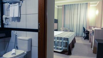 Quarto de hotel com banheiro, cama, escrivaninha e cadeira.