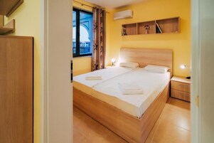 Quarto com cama de madeira, mesa de cabeceira, estante com livros e janela com cortinas.