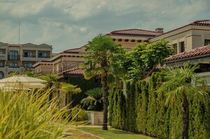 Zona residencial com várias casas, telhados em tijolos vermelhos e vegetação exuberante, incluindo palmeiras e sebes.