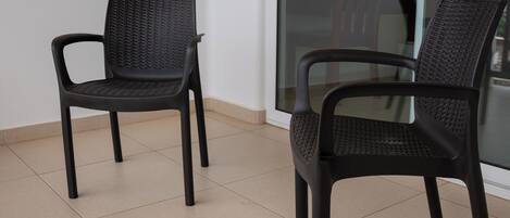 Duas cadeiras de plástico preto com assentos e encostos trançados, sobre um chão de azulejos, ao lado de uma porta de vidro que dá para uma varanda.