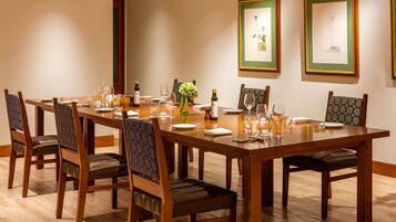 Uma sala de jantar com uma mesa de madeira comprida posta para uma refeição, rodeada por cadeiras estofadas.