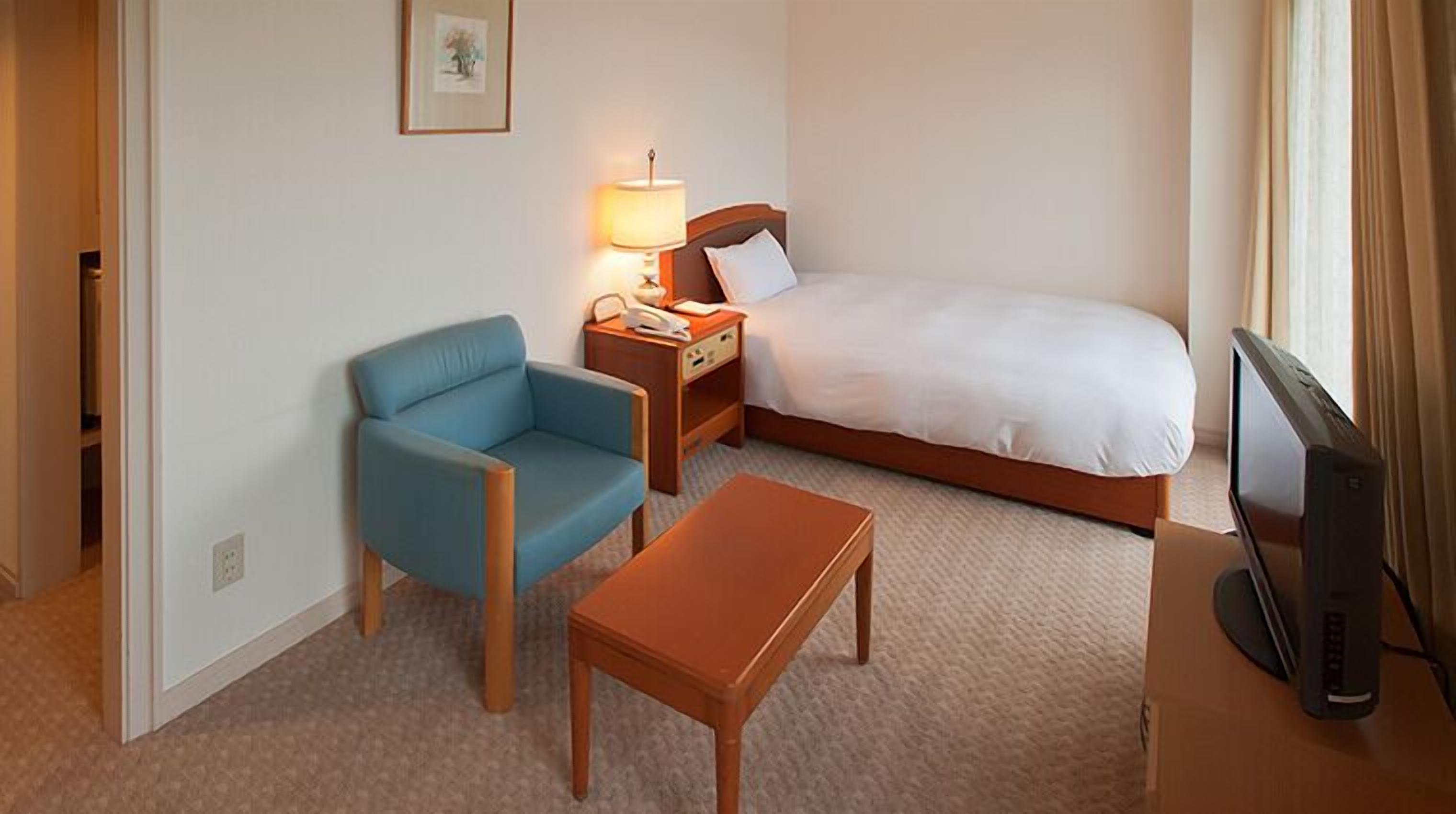 Quarto de hotel com uma cama, uma poltrona azul, uma mesinha de cabeceira de madeira com abajur, uma televisão e uma pequena mesa.