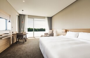 Quarto de hotel moderno com uma cama grande, uma escrivaninha com cadeira, uma mesinha com cadeira e uma janela ampla com cortinas.