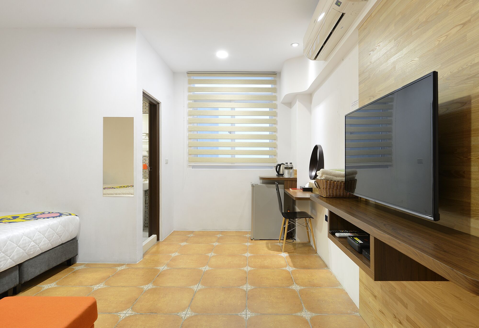Quarto compacto com cama, uma kitchenette pequena, televisão e janela com persianas.