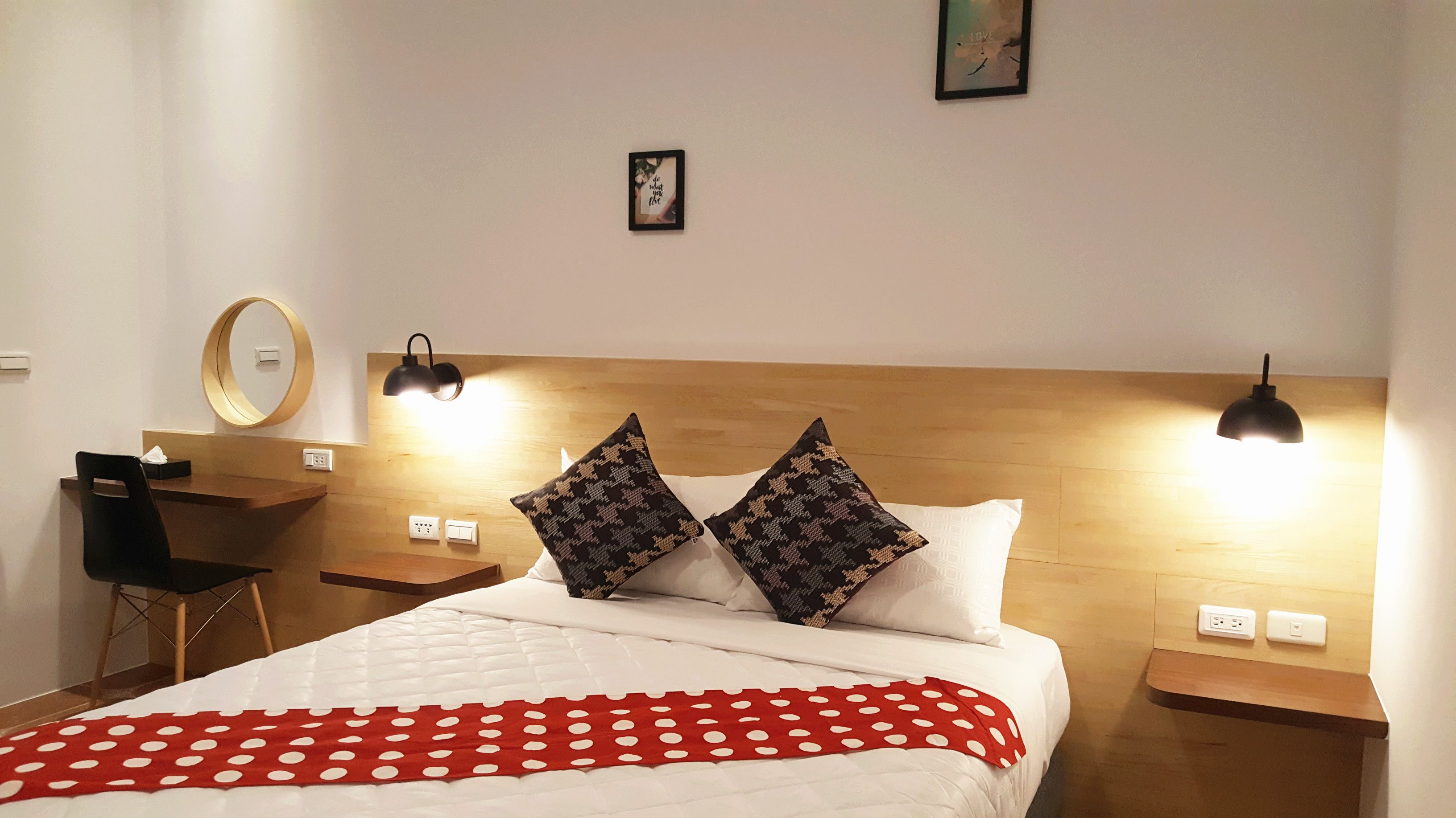 Quarto de hotel com cama, escrivaninha, cadeira, espelho redondo, duas luminárias fixadas na parede e um pequeno quadro na parede.