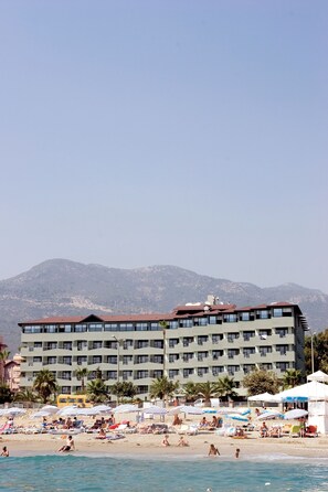 Um hotel com vários andares, uma praia em primeiro plano e montanhas ao fundo.