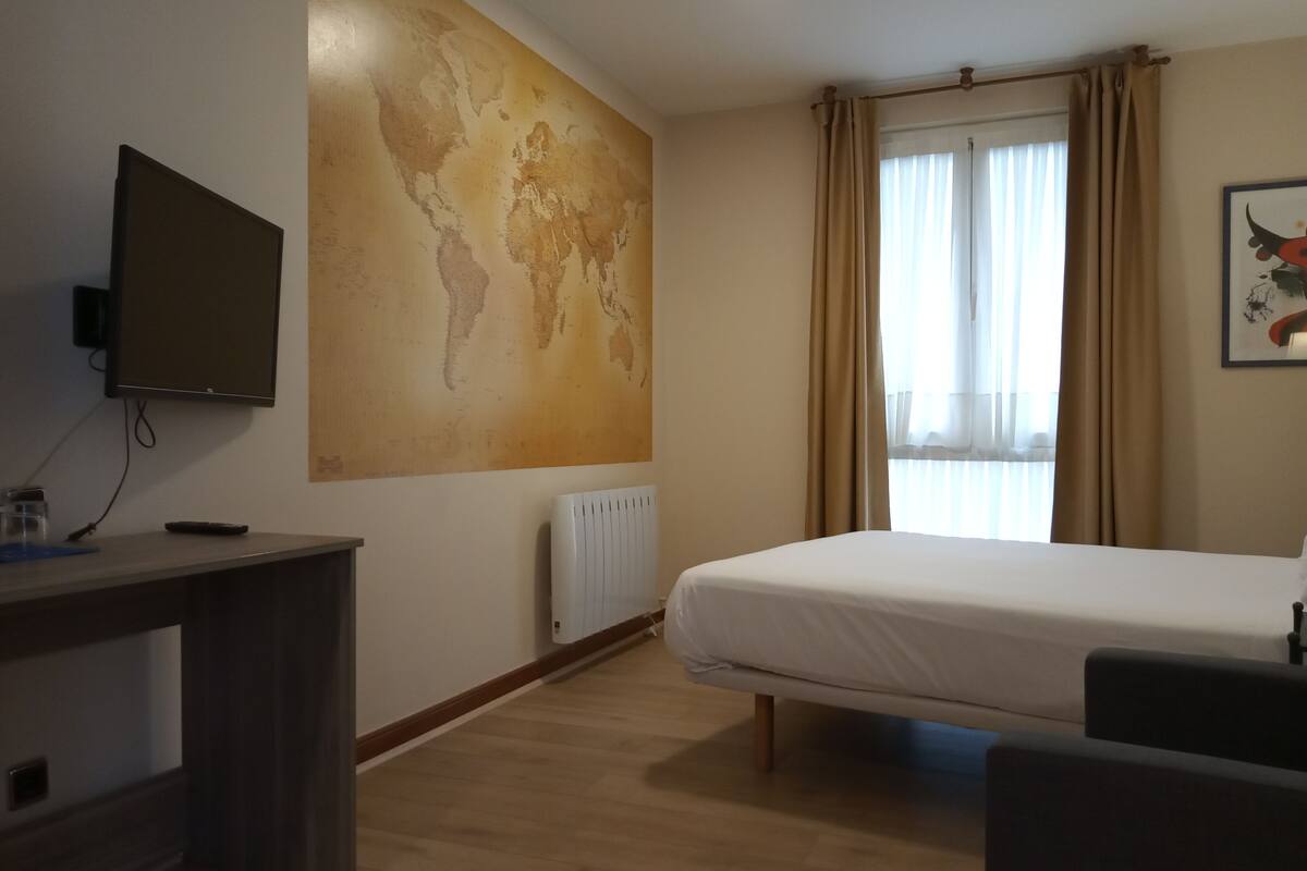 Quarto de hotel com cama, escrivaninha, mapa mundi na parede, radiador e janela com cortinas.