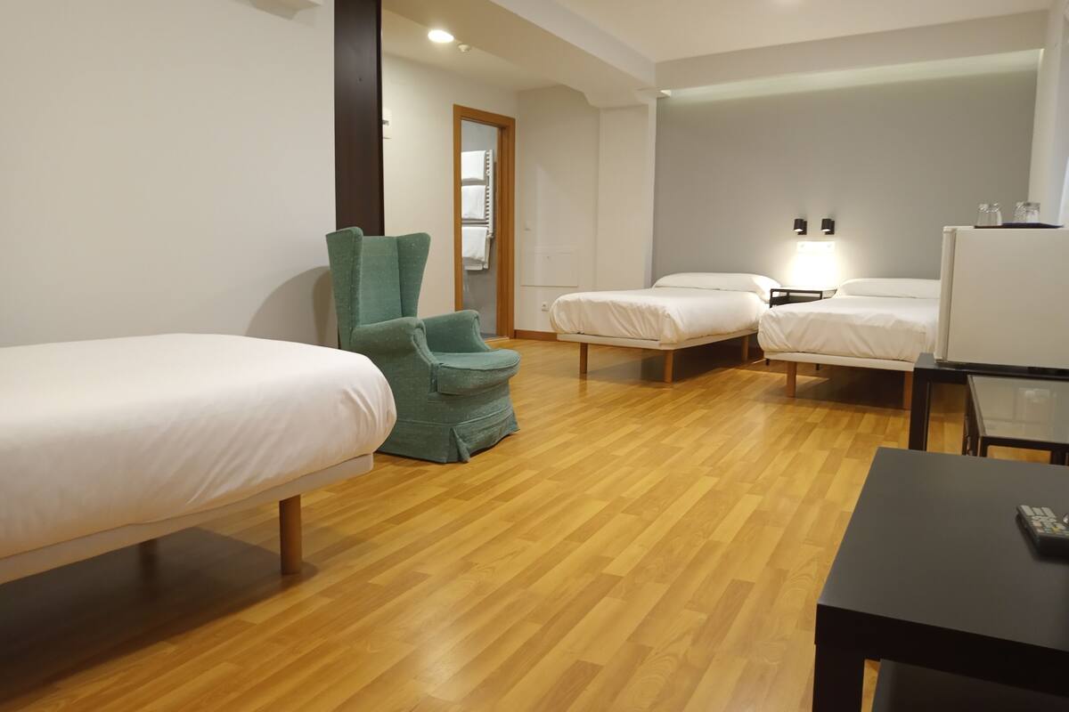 Quarto de hotel com duas camas de solteiro, uma poltrona verde, uma mesinha e um frigorífico branco.