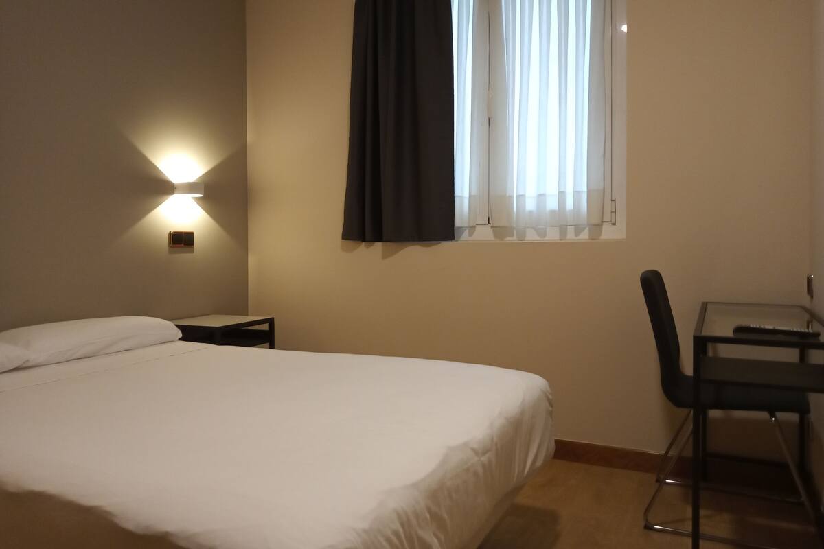 Quarto de hotel com cama, escrivaninha, cadeira, mesa de cabeceira e janela com cortinas.