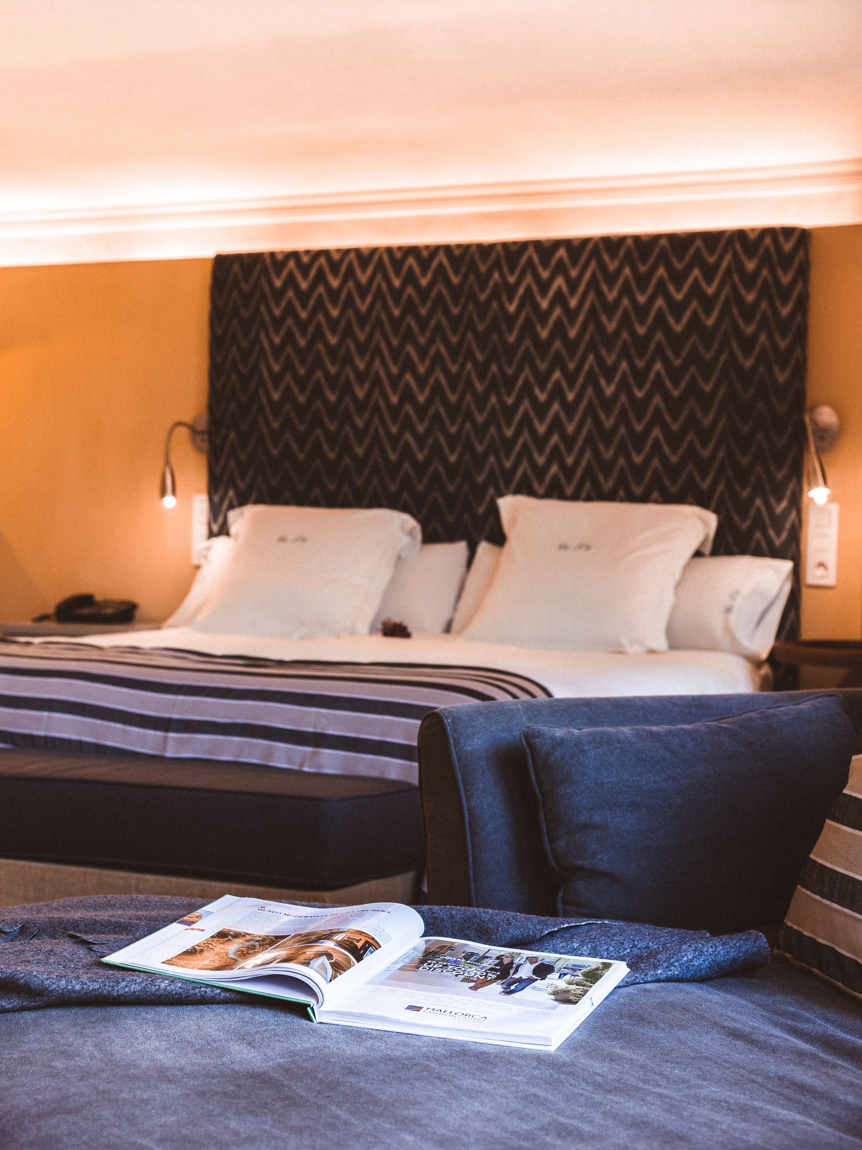 Quarto de hotel com cama, mesa de cabeceira com luminária e uma revista sobre a cama.