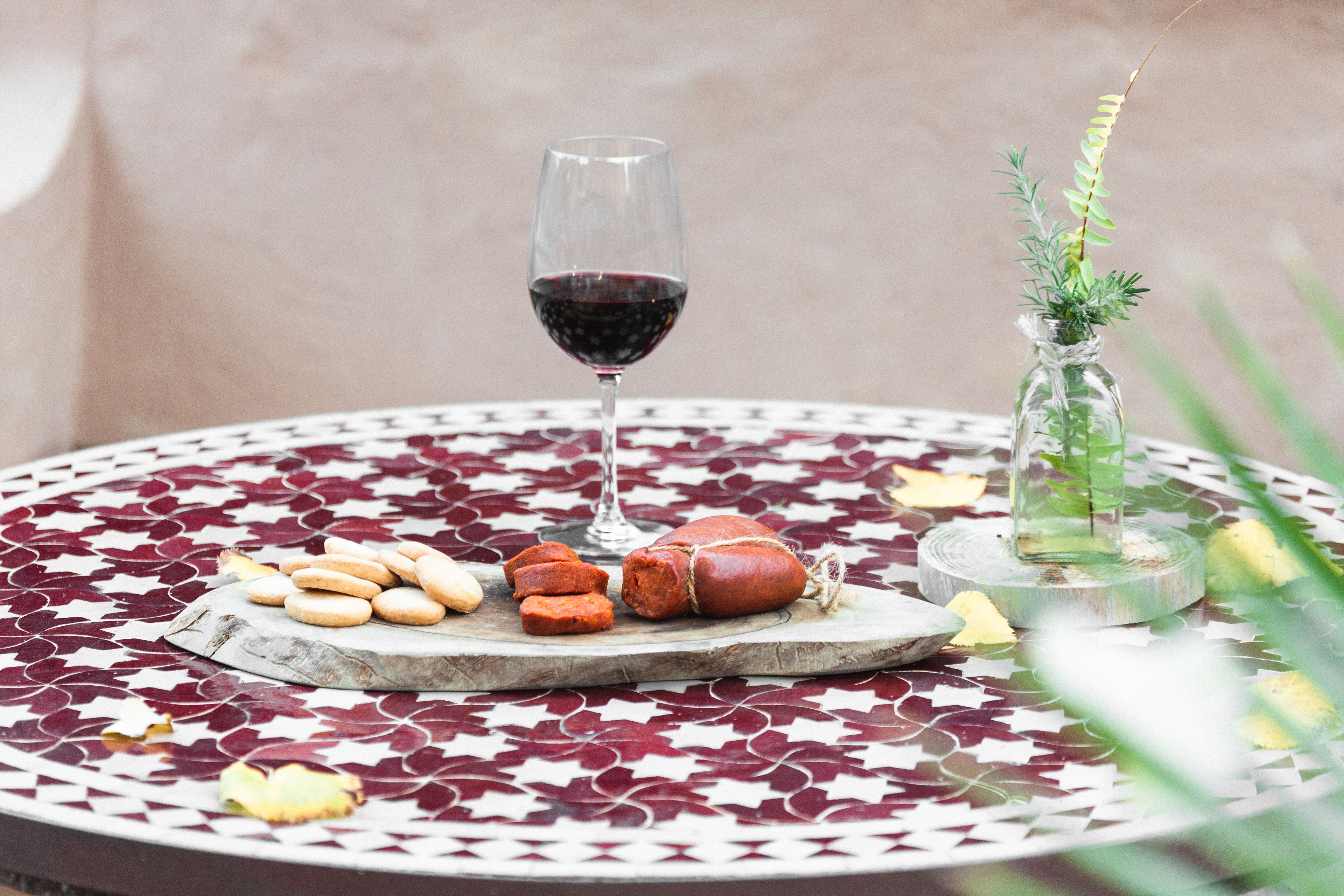 Uma mesa redonda com toalha estampada, um copo de vinho tinto, uma pequena planta em um pote de vidro e alguns alimentos.