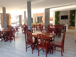 Uma ampla sala de jantar com pilares de mármore, mesas e cadeiras de madeira, e uma grande tela de televisão na parede.