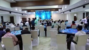 Uma sala de conferências com fileiras de cadeiras brancas, um palco com uma tela exibindo texto, e mesas com garrafas de água.