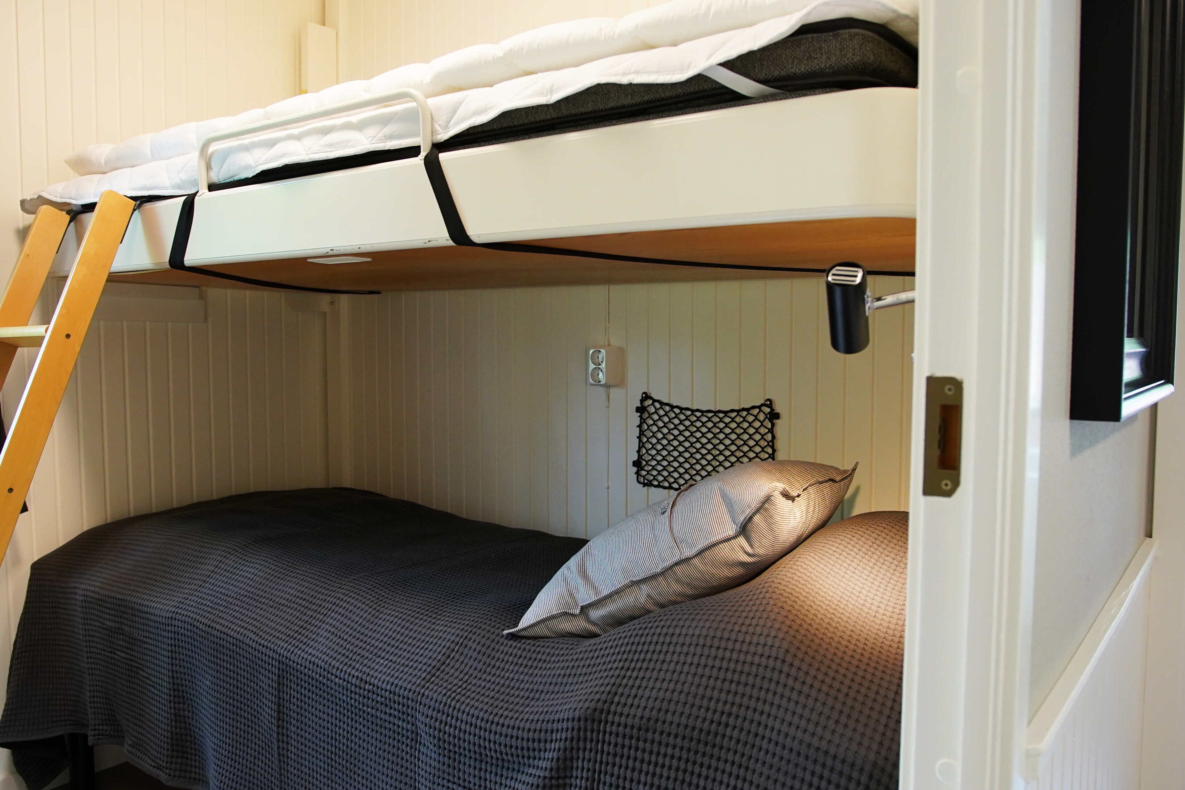 Uma cama beliche com escada, um travesseiro e um ponto de luz fixado na parede.
